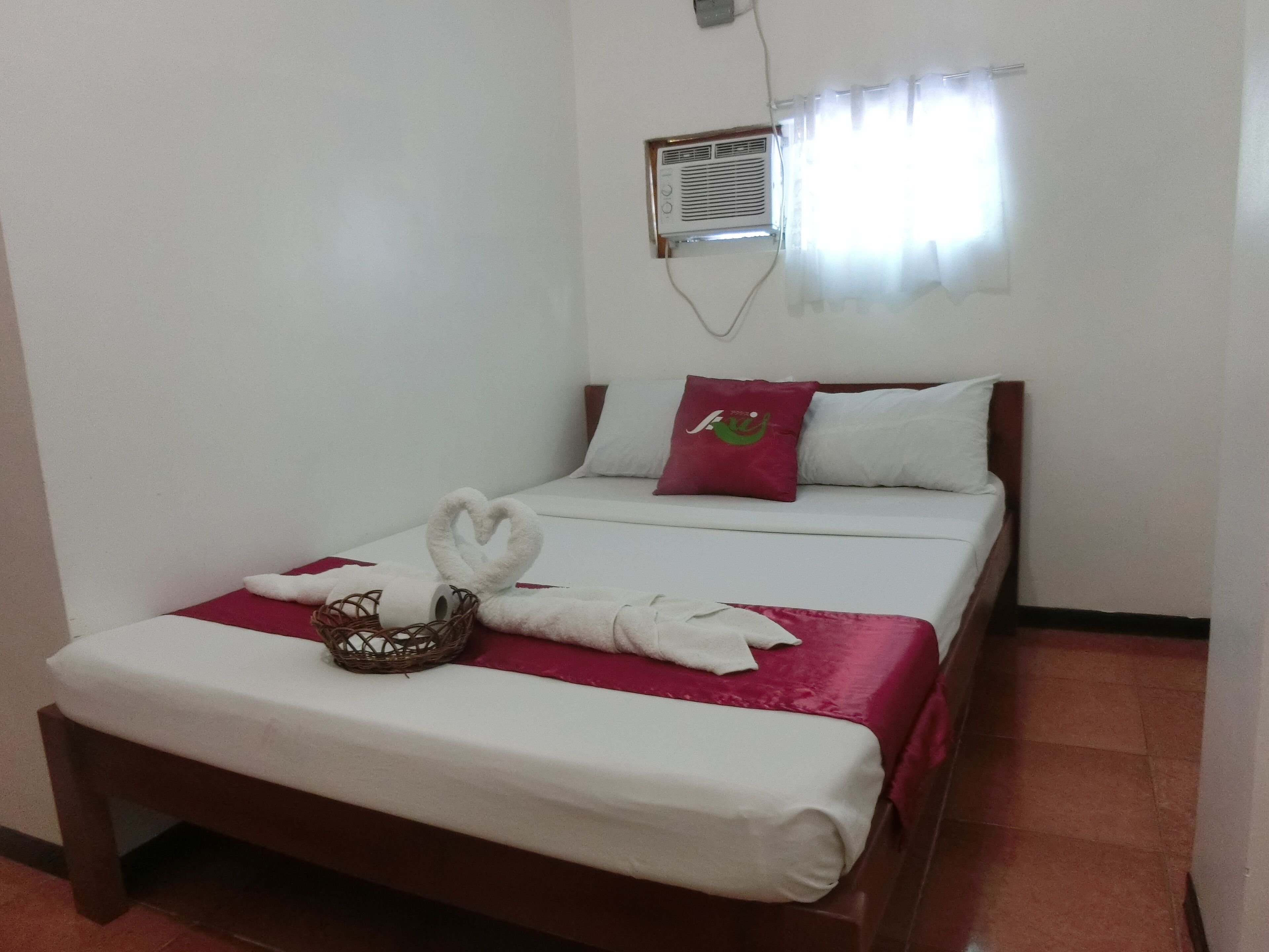 Cama arrumada com uma toalha em formato de coração, um travesseiro vermelho e uma cesta de vime.