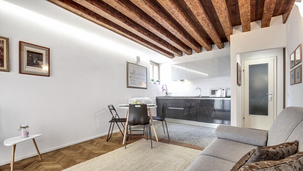 Uma sala de estar moderna com um sofá, uma mesa de jantar com cadeiras, uma cozinha ao fundo e vigas de madeira no teto.