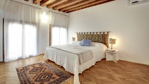 Quarto com teto de madeira, cama com colcha branca, tapete estampado e mesa de cabeceira com luminária.