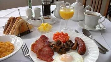 Mesa de café da manhã com ovos, salsichas, cogumelos, tomates e torradas. Há também condimentos, uma cafeteira e um copo de suco de laranja.