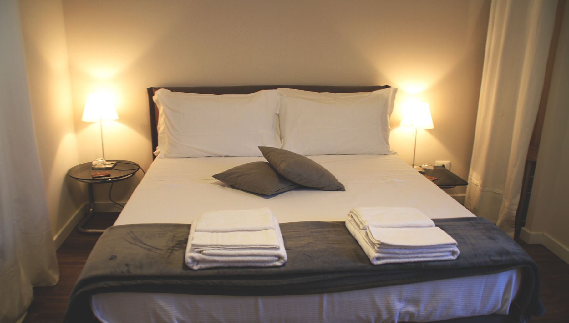 Cama bem arrumada com lençóis brancos, dois travesseiros e duas toalhas dobradas sobre um cobreleite escuro. Há duas mesas de cabeceira com abajures em ambos os lados da cama.