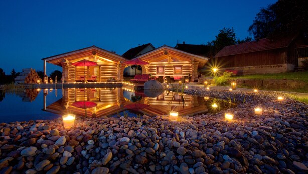 Uma cabana de toras com reflexo em um lago, rodeada por pedrinhas e iluminada por luzes.