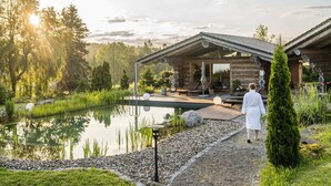 Uma cabana de tronco com um lago, rodeada por vegetação e um céu claro.