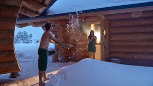 Uma cabana de madeira com exterior coberto de neve e uma pessoa jogando neve para dentro.