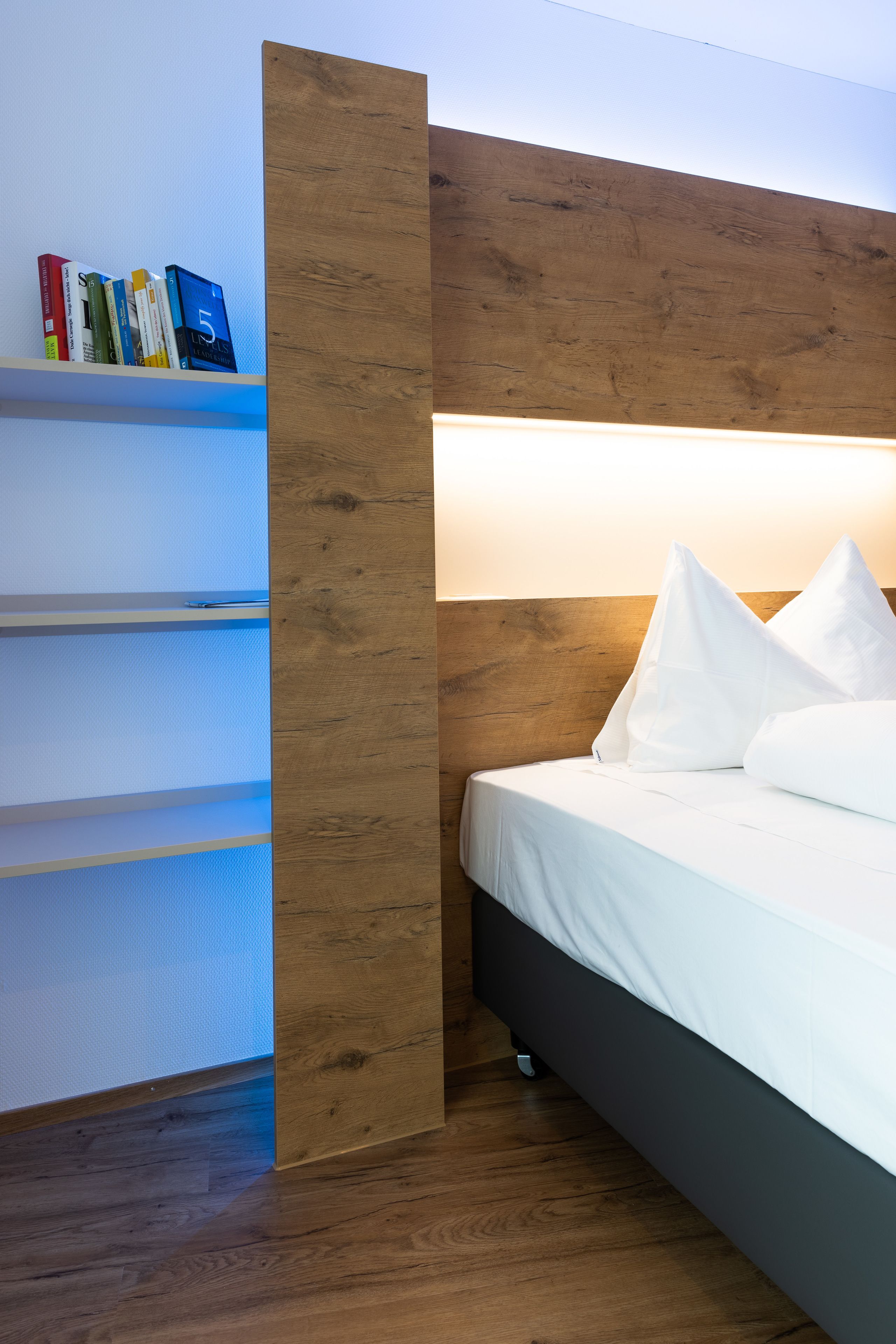 Quarto moderno com cabeceira de madeira, cama com roupa de cama branca e prateleira com livros.