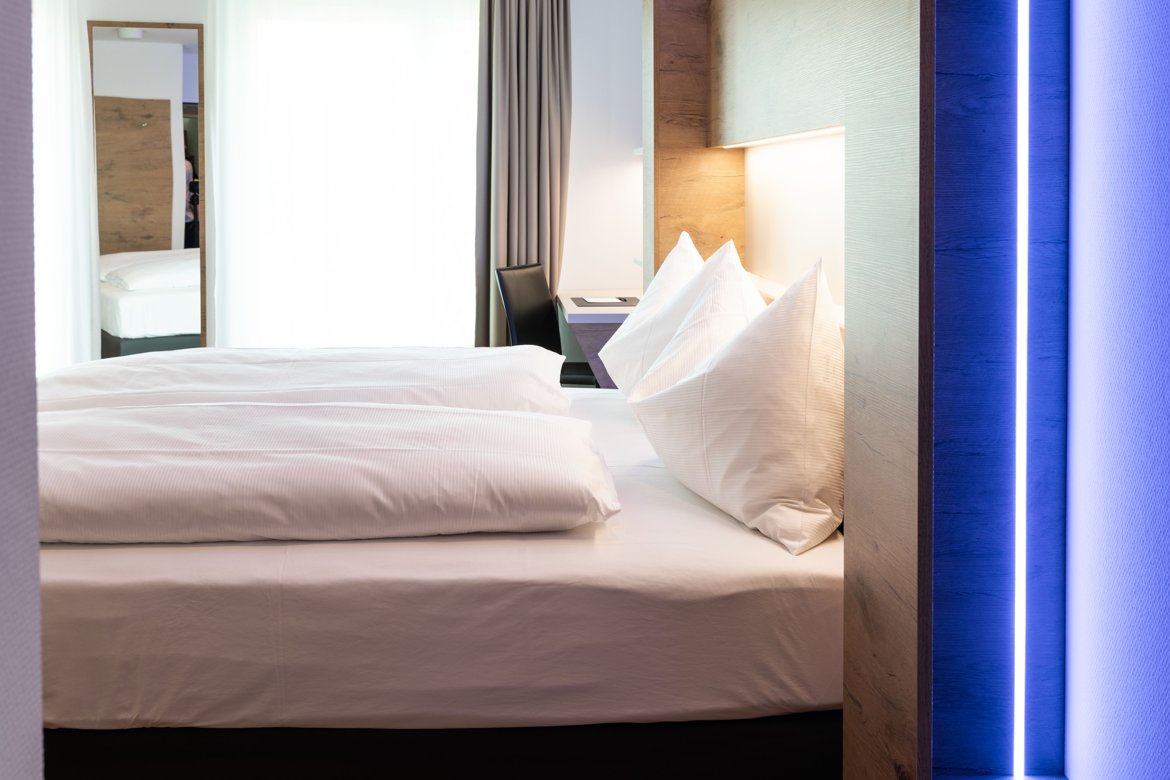 Quarto de hotel com cama bem arrumada, escrivaninha com cadeira e janela com cortinas.