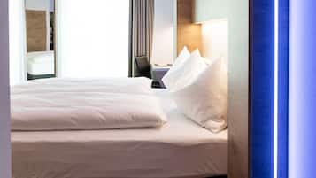 Quarto de hotel com cama bem arrumada, escrivaninha com cadeira e janela com cortinas.