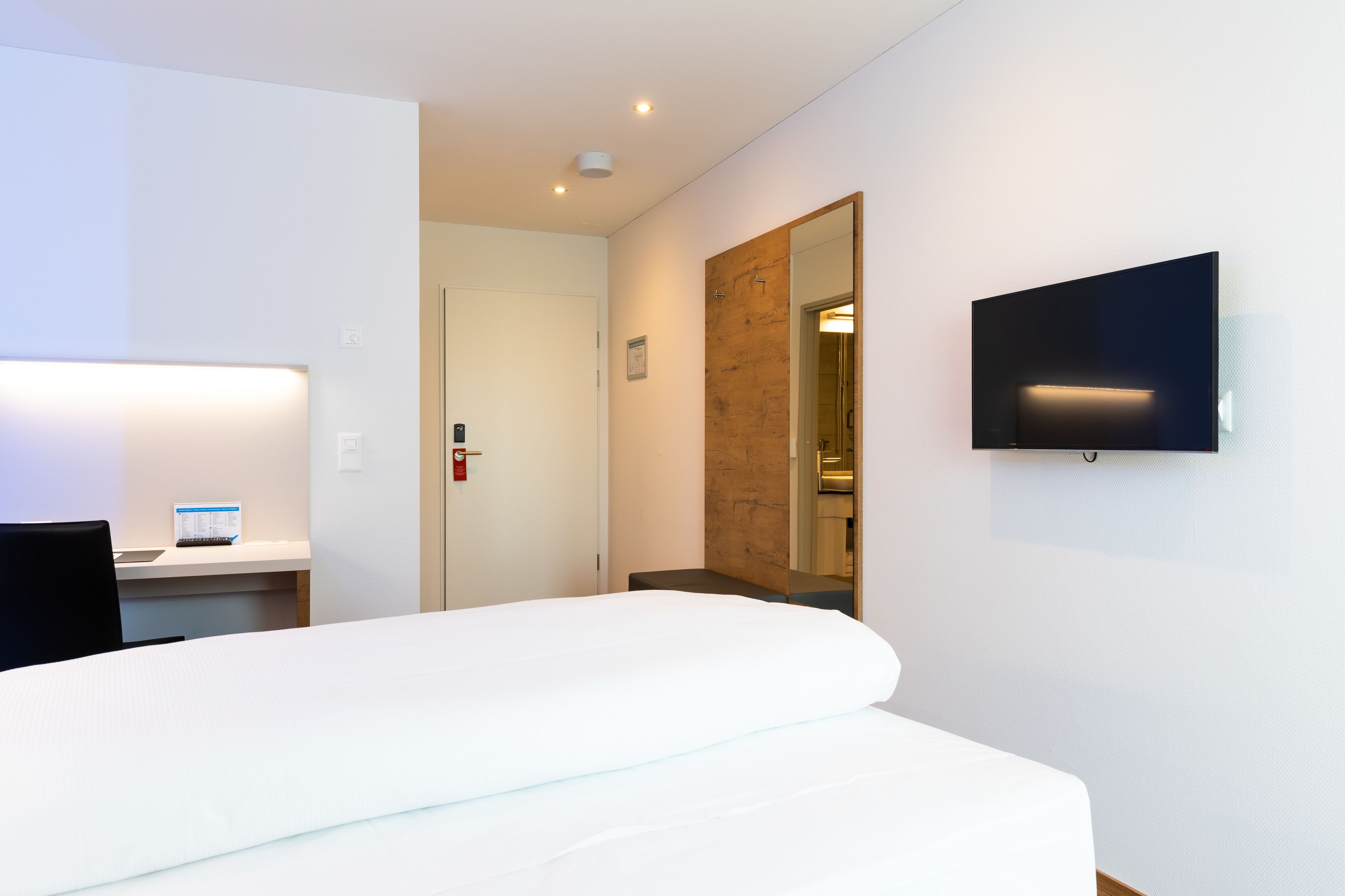 Quarto de hotel com cama, escrivaninha, televisão na parede e painel de madeira.