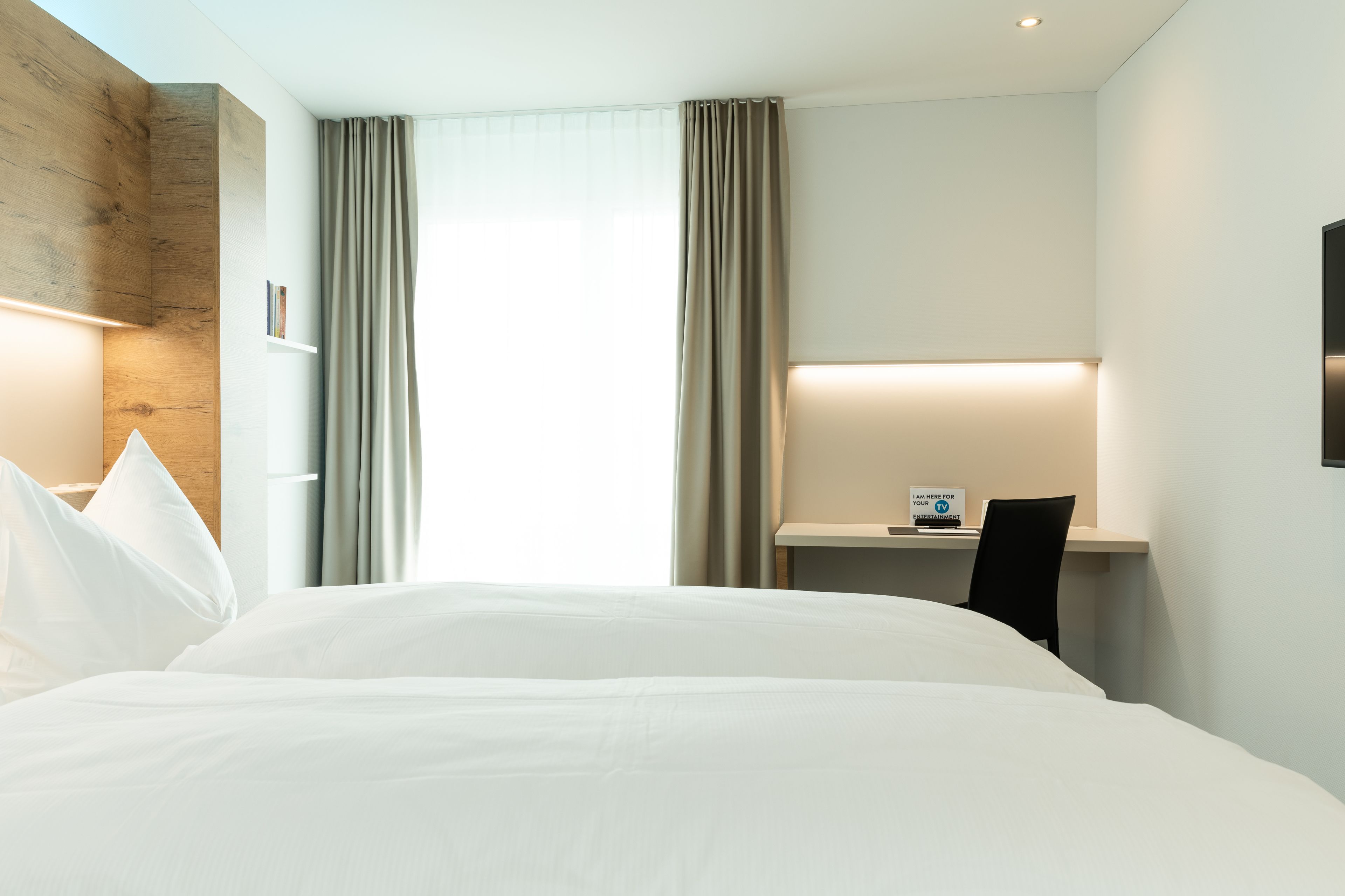 Quarto de hotel com uma cama grande, uma mesa com cadeira e uma janela com cortinas.