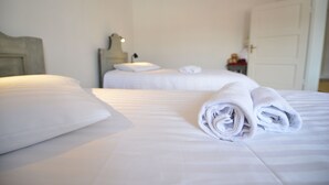 Quarto de hotel com duas camas, roupa de cama branca, mesa de cabeceira com luminária e porta fechada.