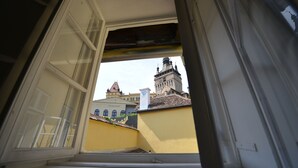 Uma janela com uma persiana aberta, mostrando vista de um edifício com torre do relógio e fachada amarela.