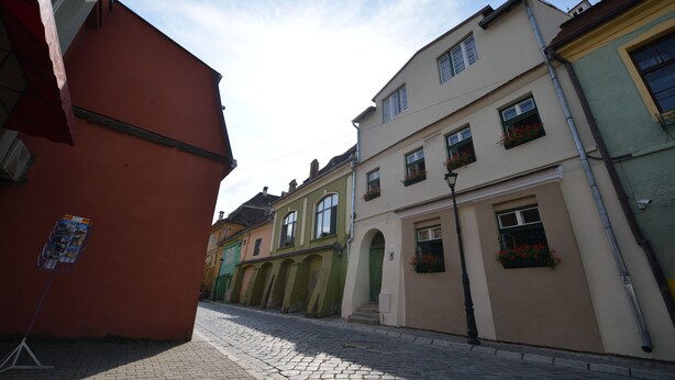 Uma rua de paralelepípedos estreita, ladeada por edifícios coloridos, com um edifício vermelho à esquerda e um edifício bege com persianas verdes à direita.