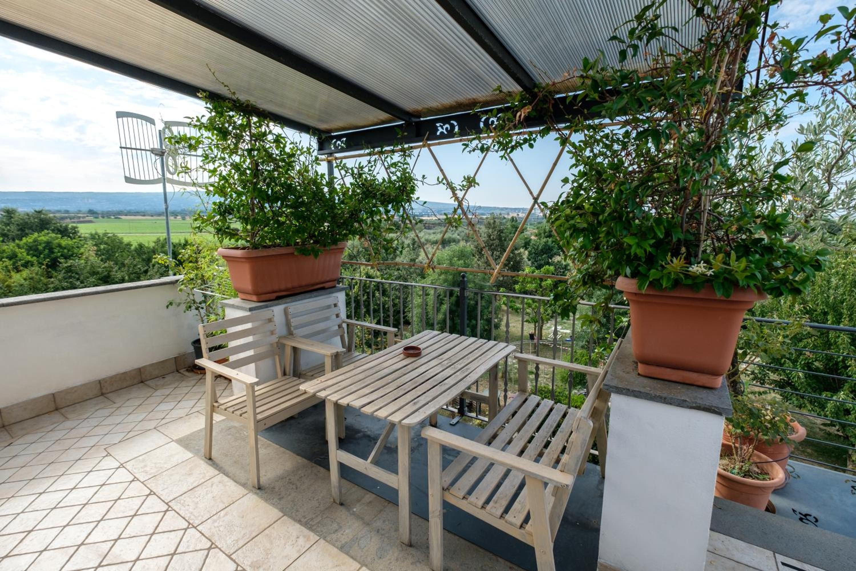 Uma varanda com mesa e cadeiras, plantas em vasos e vista para o campo.