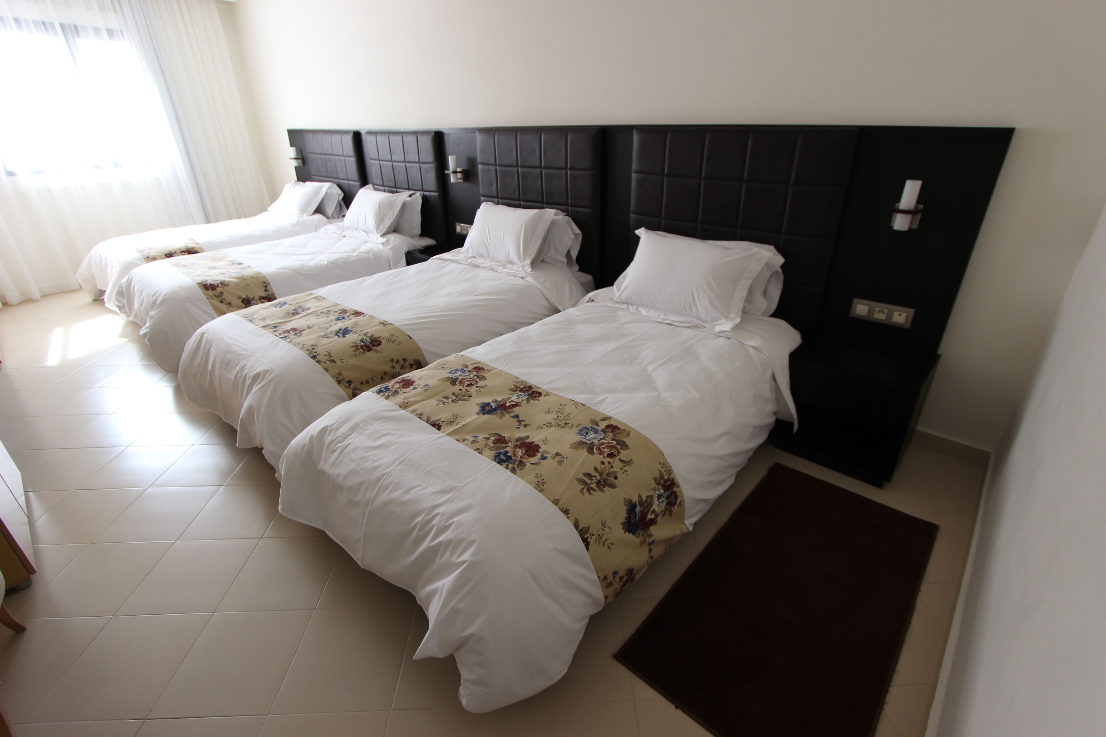 Quarto de hotel com três camas de solteiro, uma cabeceira escura e um tapete marrom.