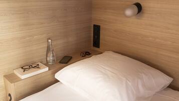 Cama bem arrumada com lençóis brancos, mesa de cabeceira de madeira com uma garrafa, copos e um smartphone.