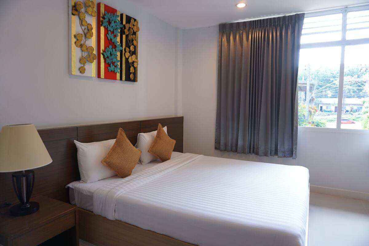 Quarto de hotel com cama bem arrumada, mesa de cabeceira com abajur e uma janela grande com cortinas.