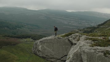 Uma pessoa em um afloramento rochoso contemplando um vale com colinas ao longe.