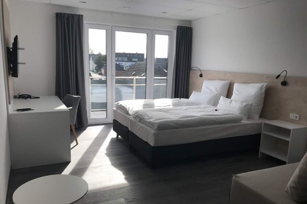 Quarto de hotel com cama, escrivaninha, cadeira, mesinha e vista para o exterior pela janela.