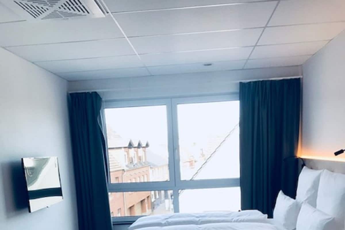 Quarto de hotel com uma cama grande, televisão de tela plana na parede e vista para edifícios pela janela.