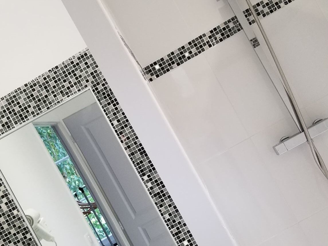 Casa de banho com parede revestida de azulejos brancos, zona de duche e espelho com moldura em mosaico preto e branco.