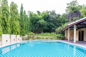 Uma piscina com padrão quadriculado, rodeada por vegetação e uma área coberta com pilares.