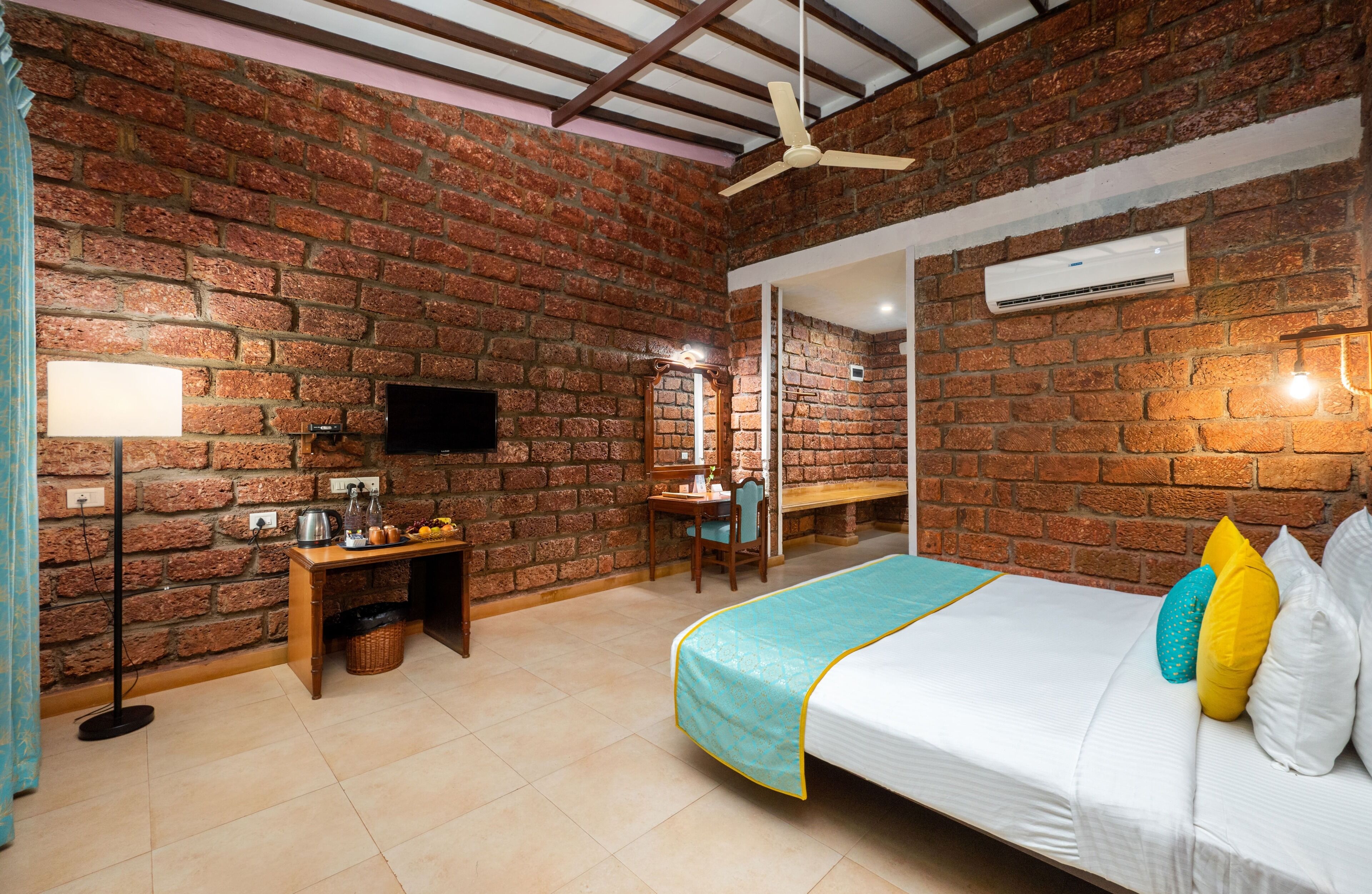 Quarto com parede de tijolos, cama com colcha azul, escrivaninha com cadeira, televisão e aparelho de ar condicionado.