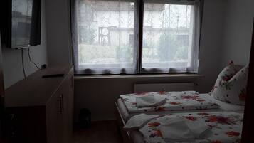 Quarto com cama, janela com cortinas, radiador, televisão fixada na parede e um pequeno armário.