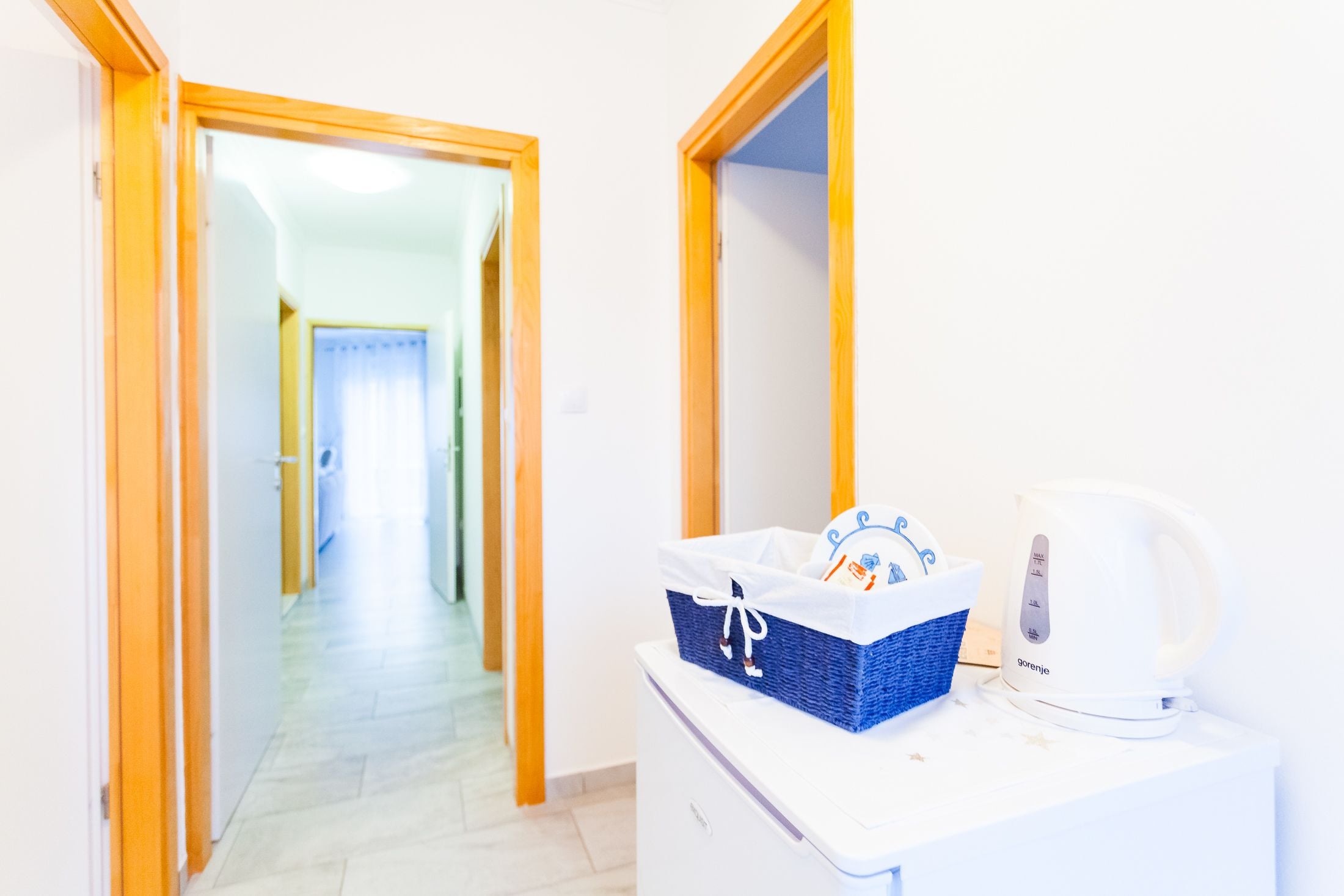 Quarto de hotel com porta de madeira, bancada branca com cesta azul e chaleira elétrica branca.
