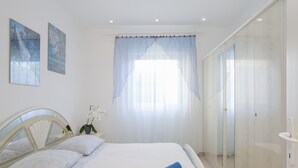 Quarto com cama, janela com cortinas leves, uma planta em vaso e um guarda-roupa.