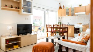 Uma sala de estar moderna com televisão, sofá e espaço para refeições. Possui cozinha com armários e janela.