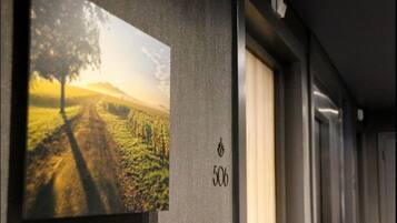 Um corredor com um quadro de paisagem emoldurado, uma porta com número e uma luminária.