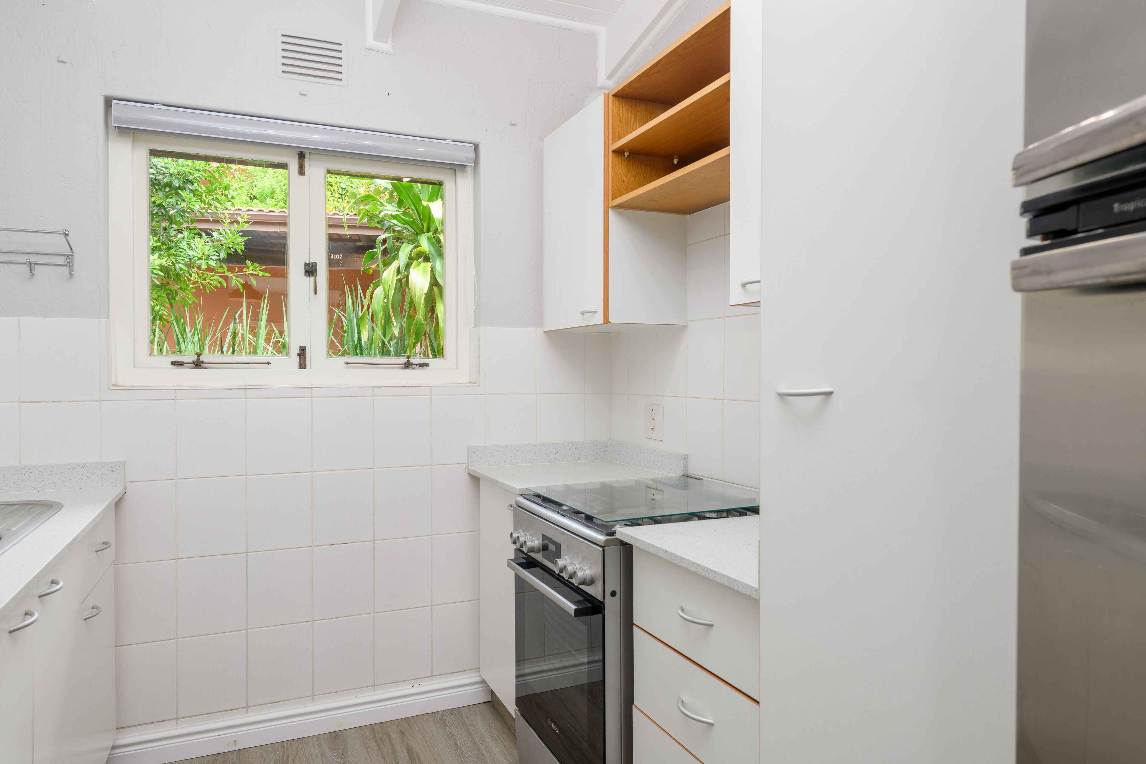 Uma cozinha compacta com armários brancos, forno embutido e janela com vista para o verde.