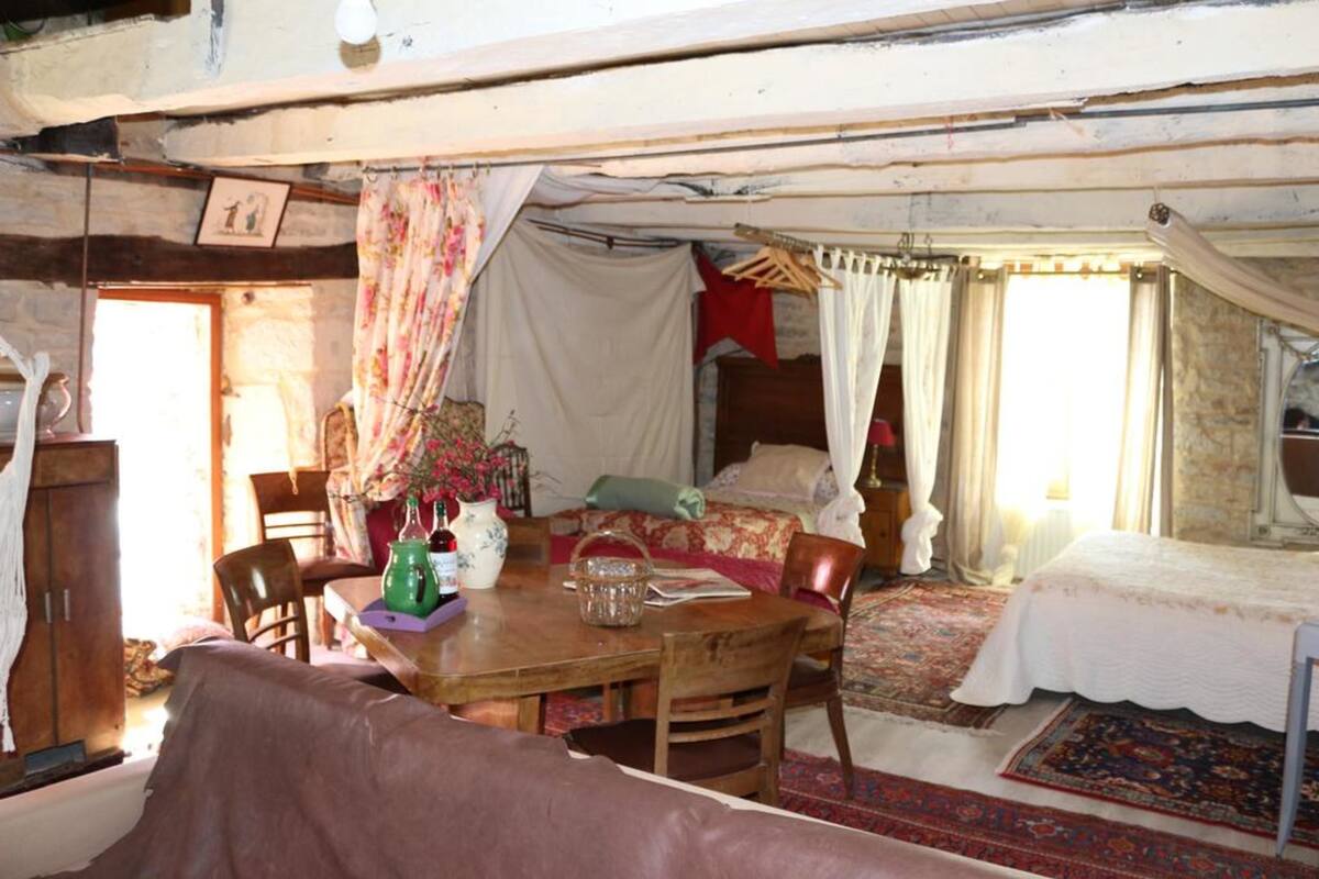 Quarto aconchegante com mesa de jantar de madeira, cadeiras, uma cama com colcha branca e uma janela grande com cortinas.