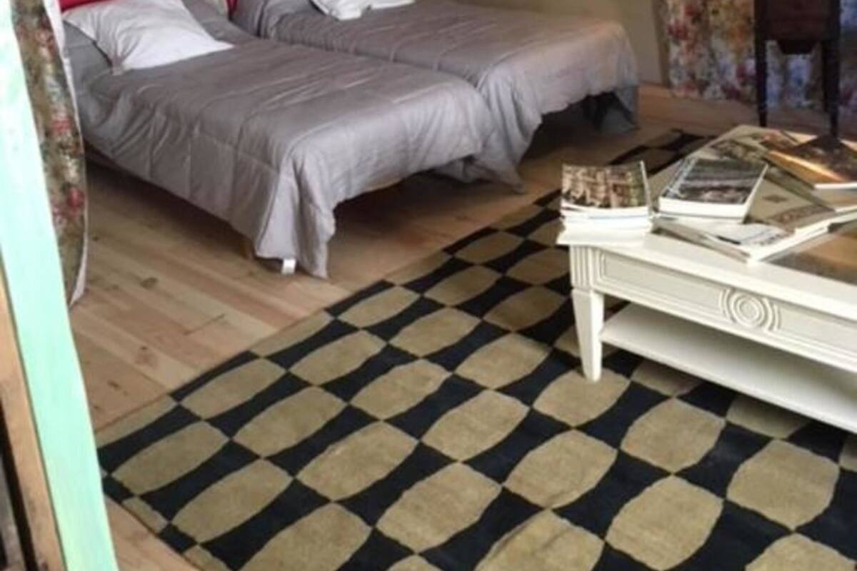 Quarto com duas camas, papel de parede floral, tapete xadrez e uma mesa com livros.