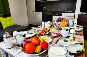 Mesa de café da manhã posta com pratos de fruta, ovos, torradas e suco em uma cozinha moderna, com um sofá e cortinas verdes ao fundo.