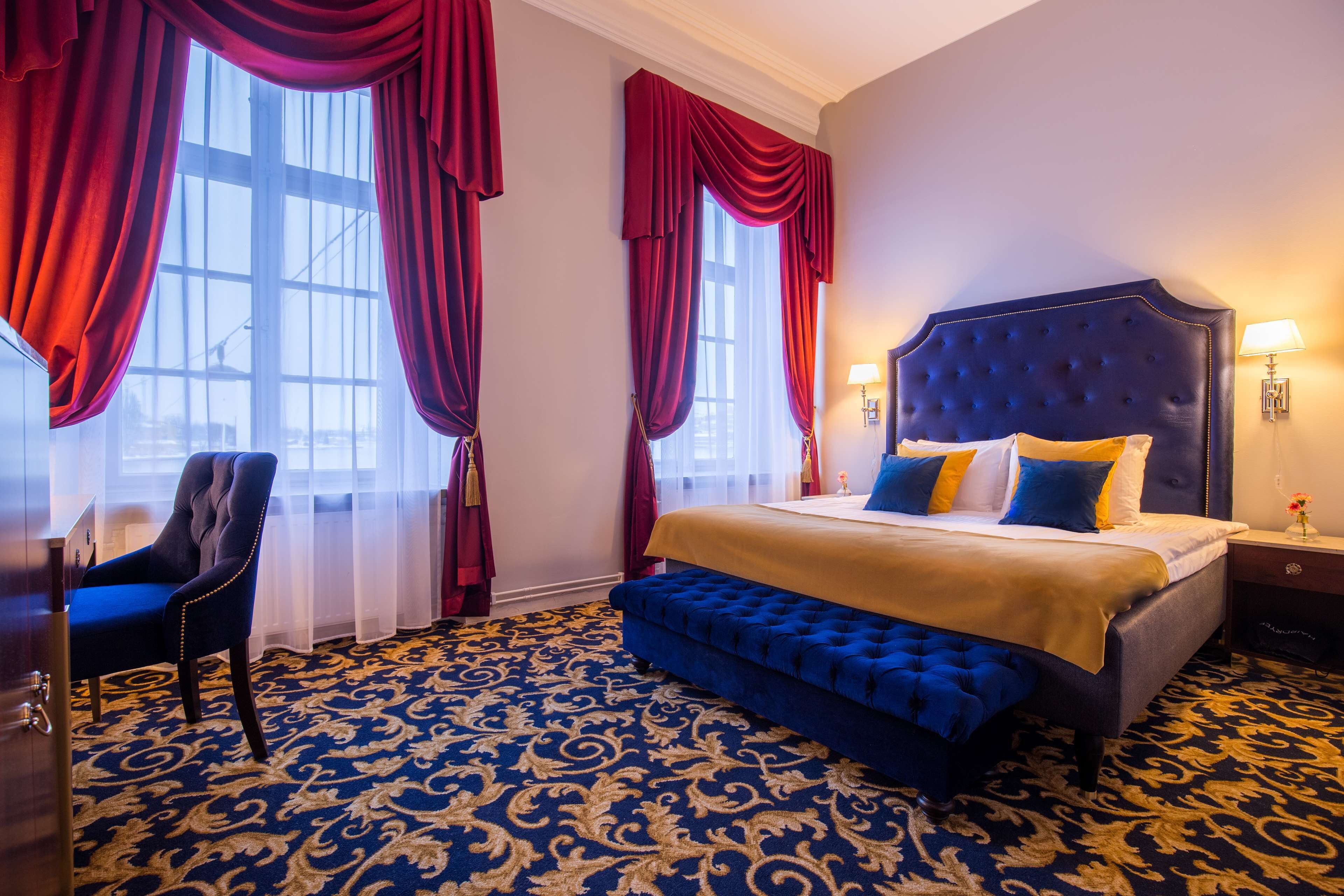 Quarto de hotel com uma cama grande, uma poltrona estofada em azul e um tapete estampado.