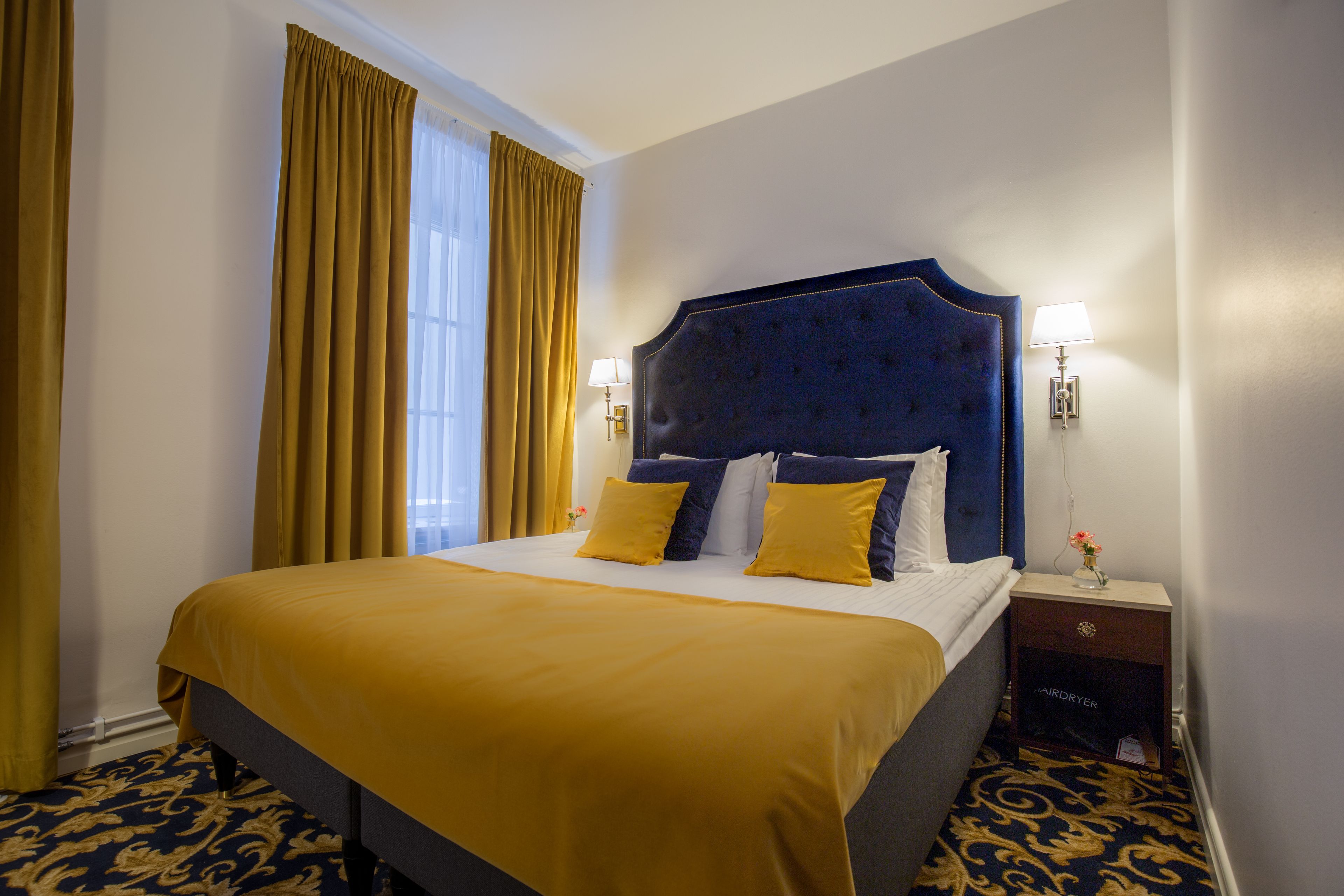 Quarto de hotel com uma cama grande, cortinas amarelas, cabeceira escura e mesa de cabeceira com luminária e secador de cabelo.