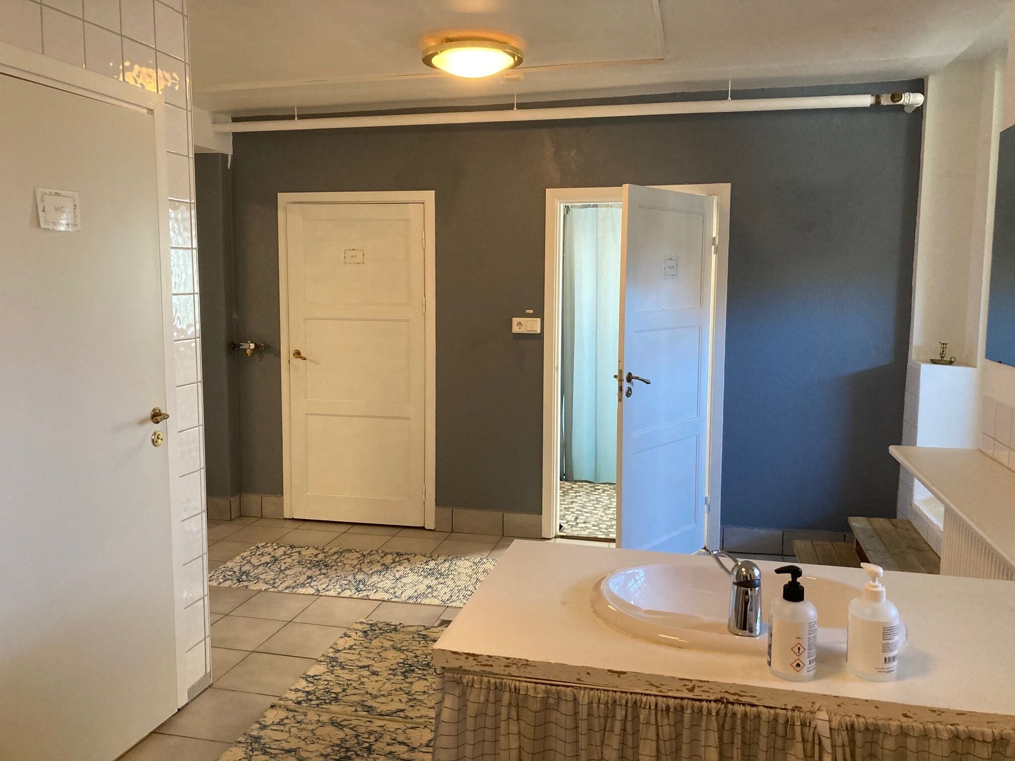 Casa de banho com lavatório branco, duas embalagens de sabonete, um espelho e uma porta com placa a indicar a casa de banho.