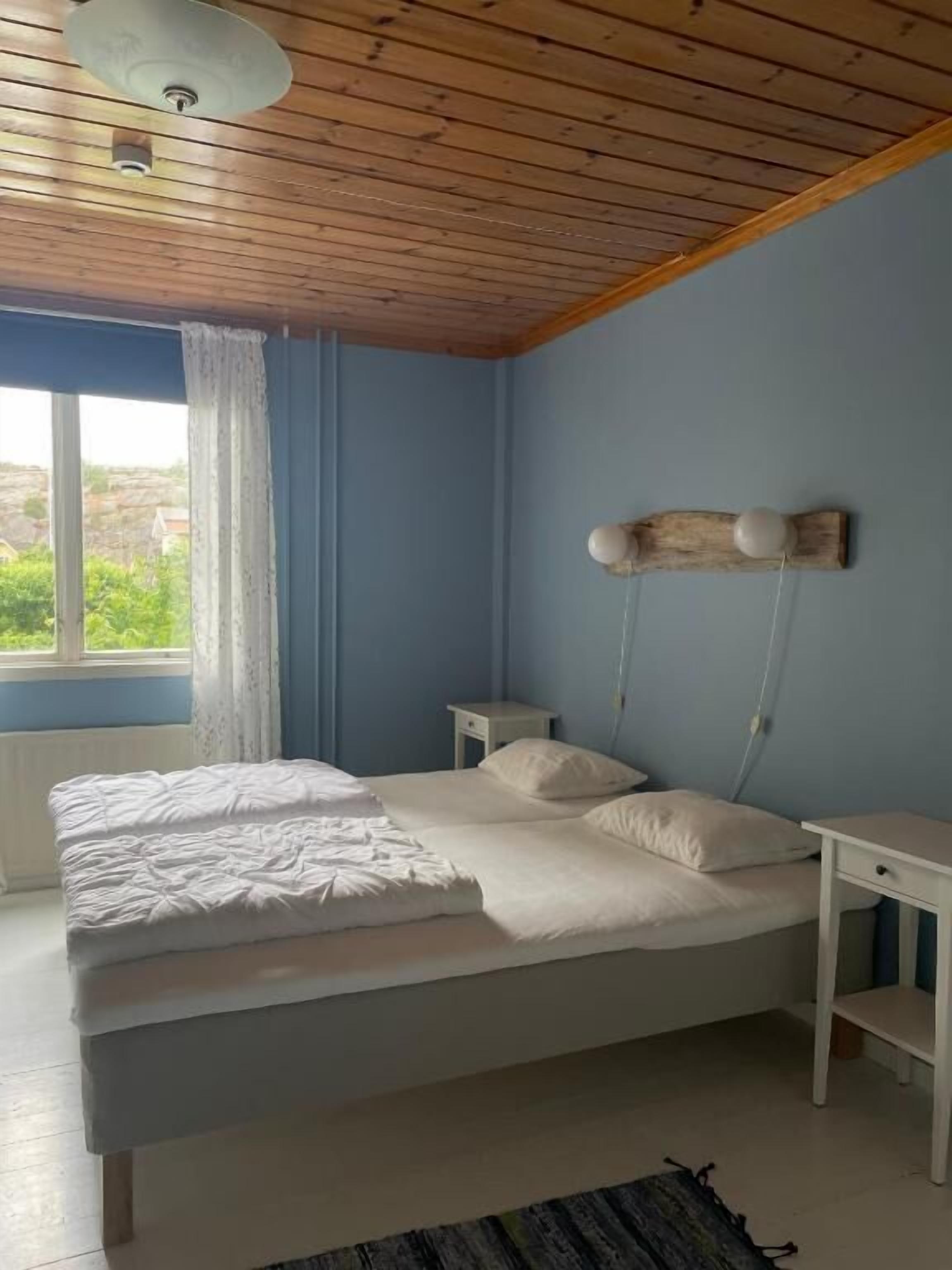 Quarto com teto de madeira, cama com roupa de cama branca, mesa de cabeceira e luminária fixada na parede.