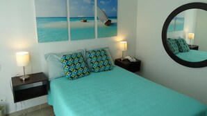 Quarto com cama, duas mesas de cabeceira, um criado-mudo, um espelho e três quadros na parede retratando uma cena de praia.