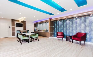 Uma área de jantar moderna com piso de madeira, uma mesa central com cadeiras verdes e cadeiras vermelhas em frente a uma parede com um painel decorativo.