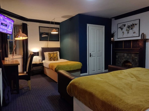 Quarto de hotel pequeno, com uma cama, escrivaninha, cadeira, mesa, abajur, televisão, porta e lareira.