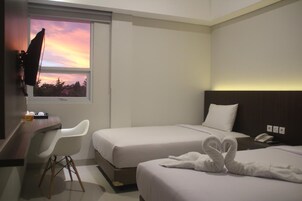 Quarto de hotel com duas camas, uma escrivaninha com cadeira, televisão na parede e vista do pôr do sol pela janela.