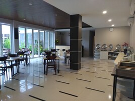 Um lobby de hotel moderno com piso revestido e limpo e um pilar central. Há mesas e cadeiras dispostas para refeições, e uma área de cozinha com eletrodomésticos e louças à vista.