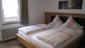Quarto com cama de madeira, duas almofadas, um cobertor, radiador, janela com cortinas e um quadro na parede.