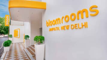 Entrada de um hotel moderno com piso xadrez, uma placa grande com a inscrição "bloomrooms Janpath, Nova Delhi" e um toldo amarelo.
