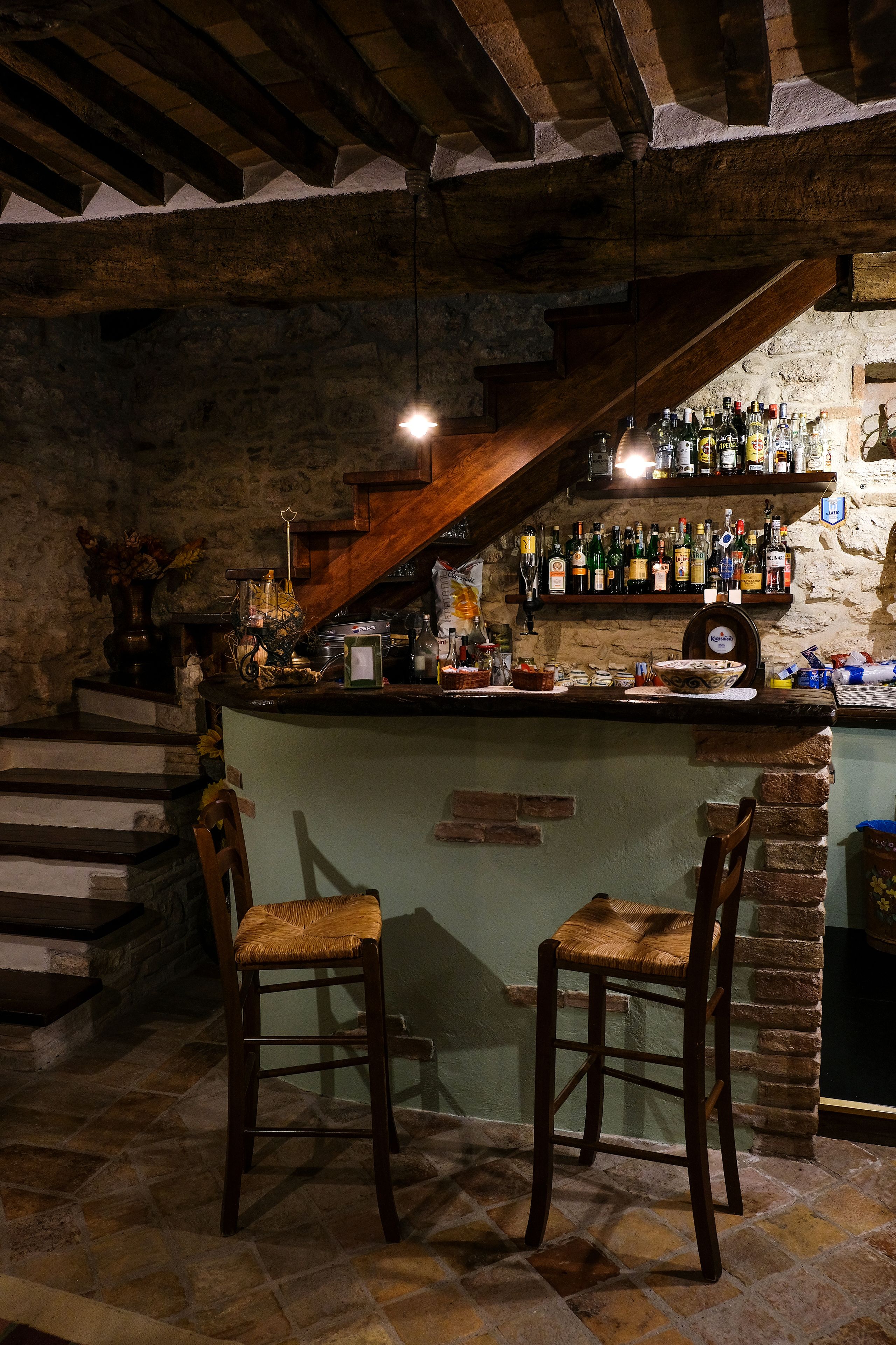 Uma área de bar com cadeiras de madeira, piso de pedra e uma escada que leva a um mezanino.
