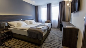 Quarto de hotel com uma cama grande, escrivaninha, cadeira, TV de tela plana e uma janela ampla com cortinas.