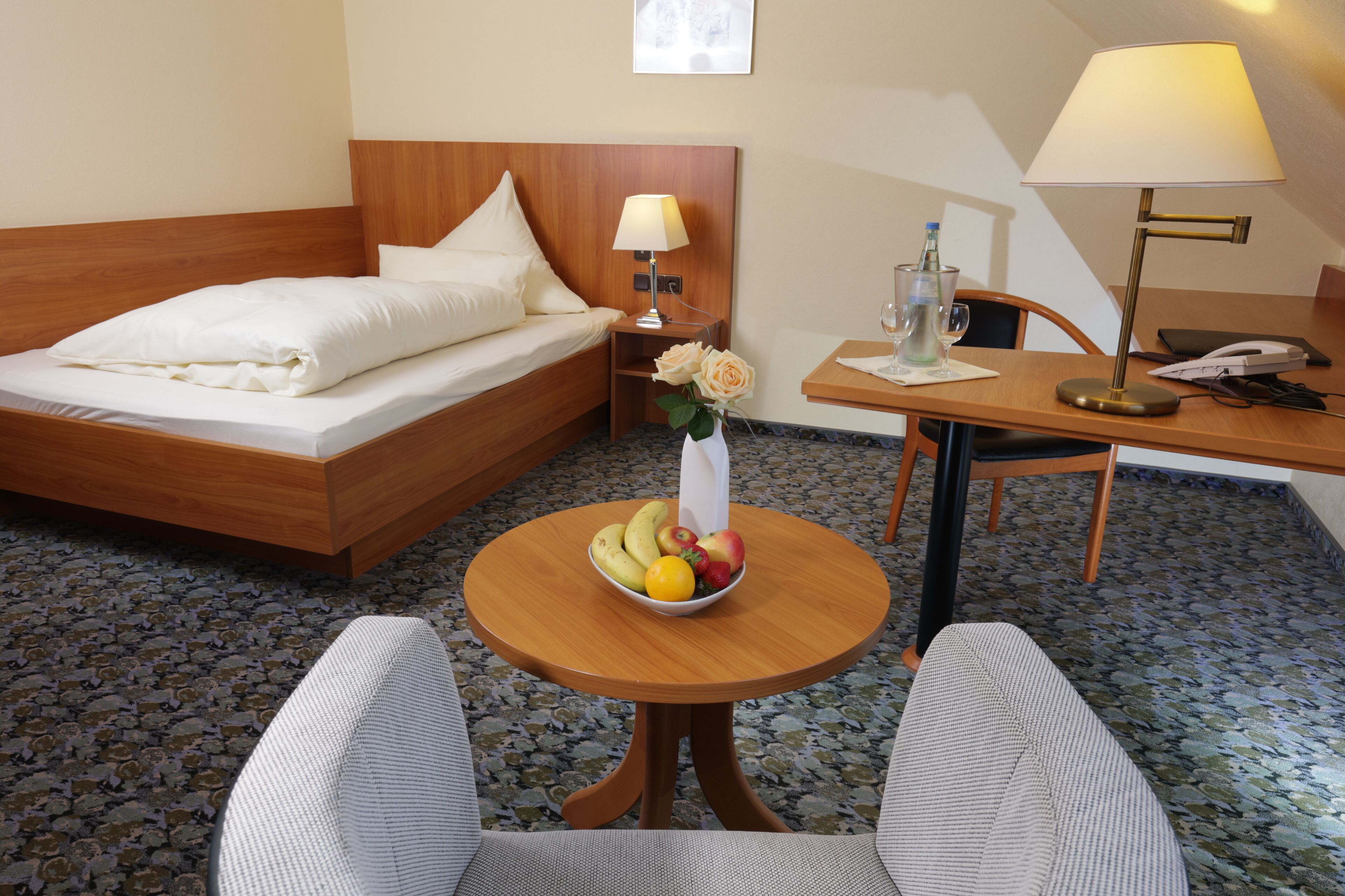 Quarto de hotel com uma cama de madeira, uma mesinha redonda com frutas, uma cadeira, uma escrivaninha com abajur e uma mesa de cabeceira com abajur.