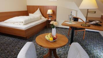 Quarto de hotel com uma cama de madeira, uma mesinha redonda com frutas, uma cadeira, uma escrivaninha com abajur e uma mesa de cabeceira com abajur.